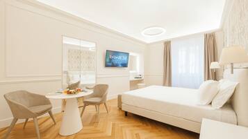 Quarto de hotel com uma cama, duas cadeiras, uma mesinha com uma bebida, uma televisão e uma janela com cortinas.