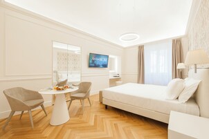 Quarto de hotel com uma cama, duas cadeiras, uma mesinha com uma bebida, uma televisão e uma janela com cortinas.