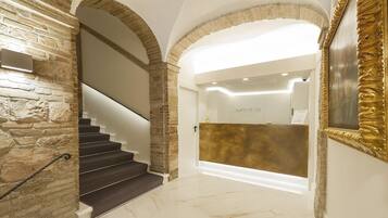 Um hall de hotel com piso de mármore, escada com degraus escuros e balcão de recepção com acabamento dourado.