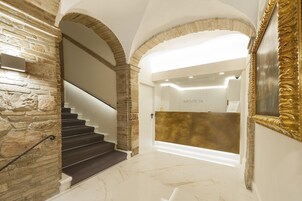 Um hall de hotel com piso de mármore, escada com degraus escuros e balcão de recepção com acabamento dourado.