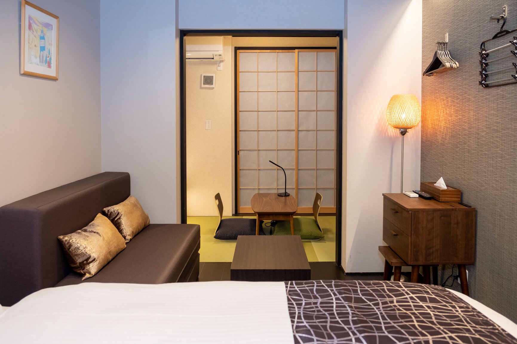 Quarto de hotel com cama, sofá, pequena mesa, escrivaninha, cadeira, abajur e porta deslizante que dá para um quarto japonês tradicional.