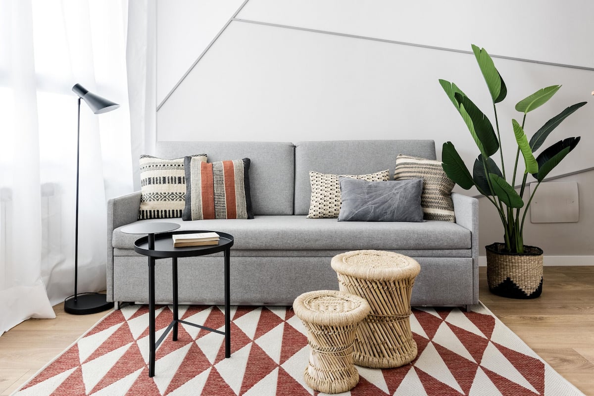 Uma sala moderna com um sofá cinzento, uma mesinha lateral redonda preta, dois taburetes trançados, uma planta verde grande e um tapete com padrão vermelho e branco.
