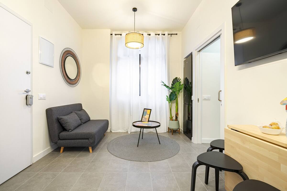 Uma sala de estar moderna com um sofá cinzento, uma mesa redonda pequena com um porta-retrato, uma planta em vaso e um espaço de jantar em madeira.