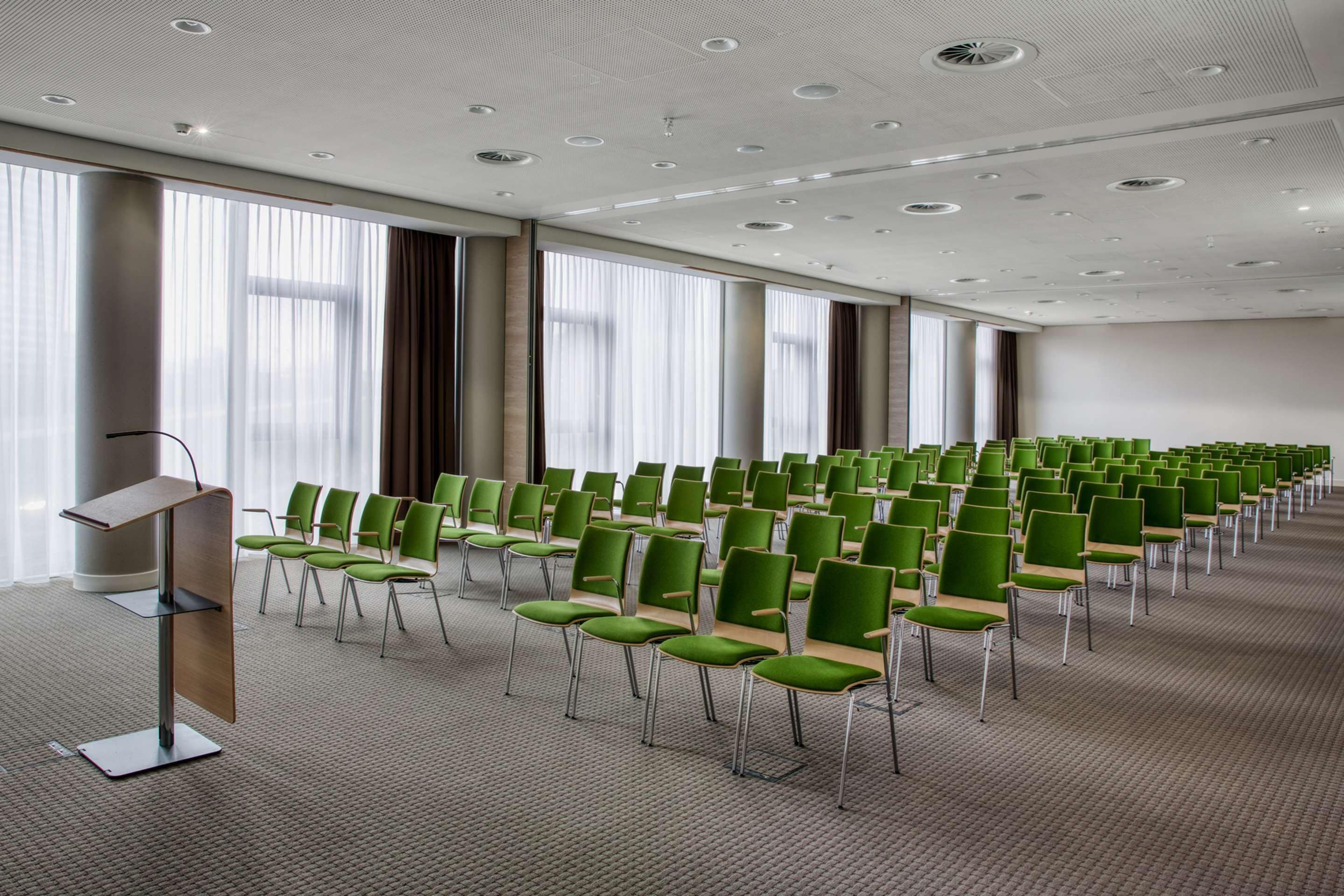 Uma sala de conferências com fileiras de cadeiras verdes, um púlpito e janelas amplas com cortinas.