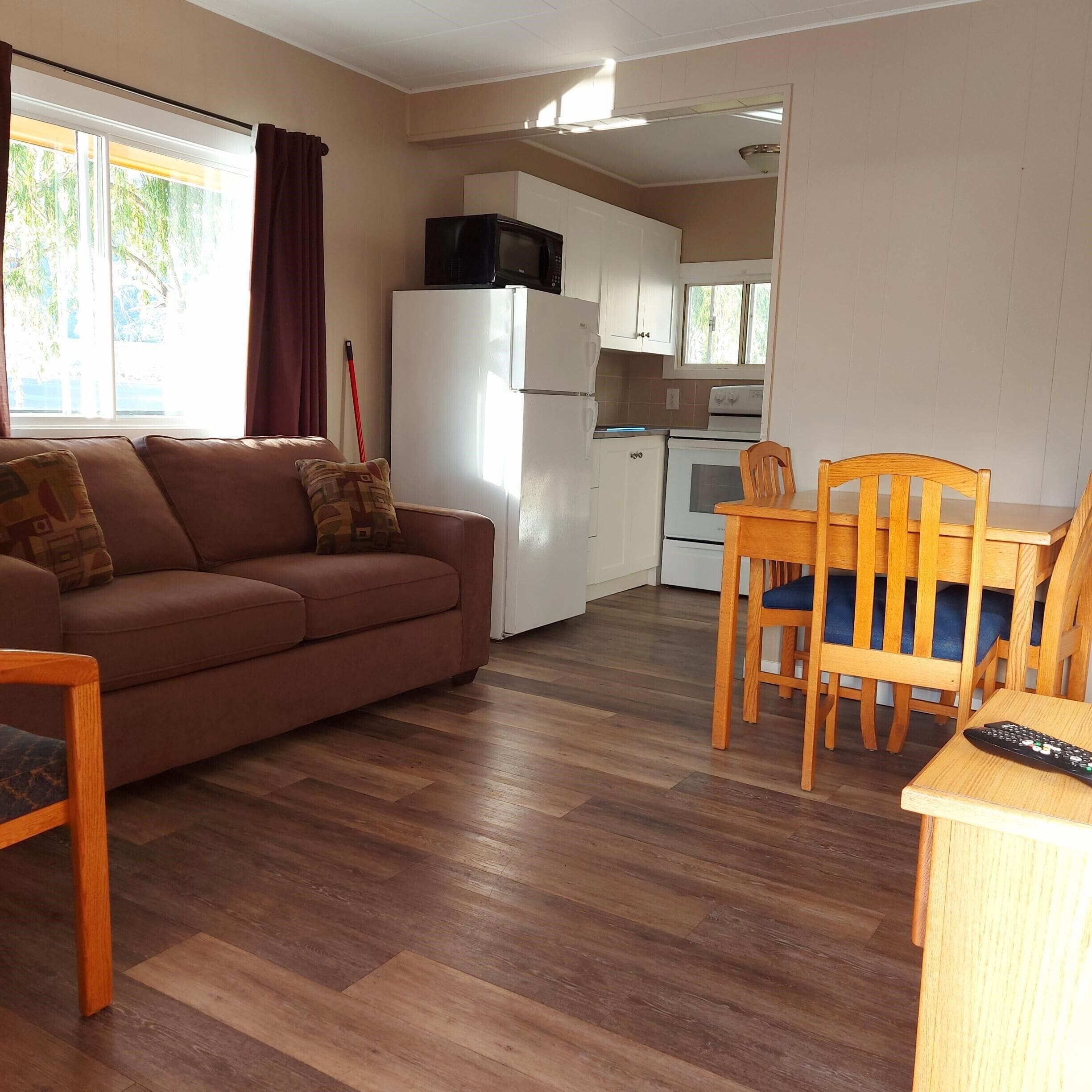 Um espaço de estar compacto com sofá, mesa de jantar, cadeiras, área de cozinha com geladeira e fogão, e janela com cortinas.