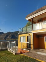 Um edifício de dois andares com uma varanda, uma porta com a inscrição "100" e vista para um lago e montanhas ao fundo.
