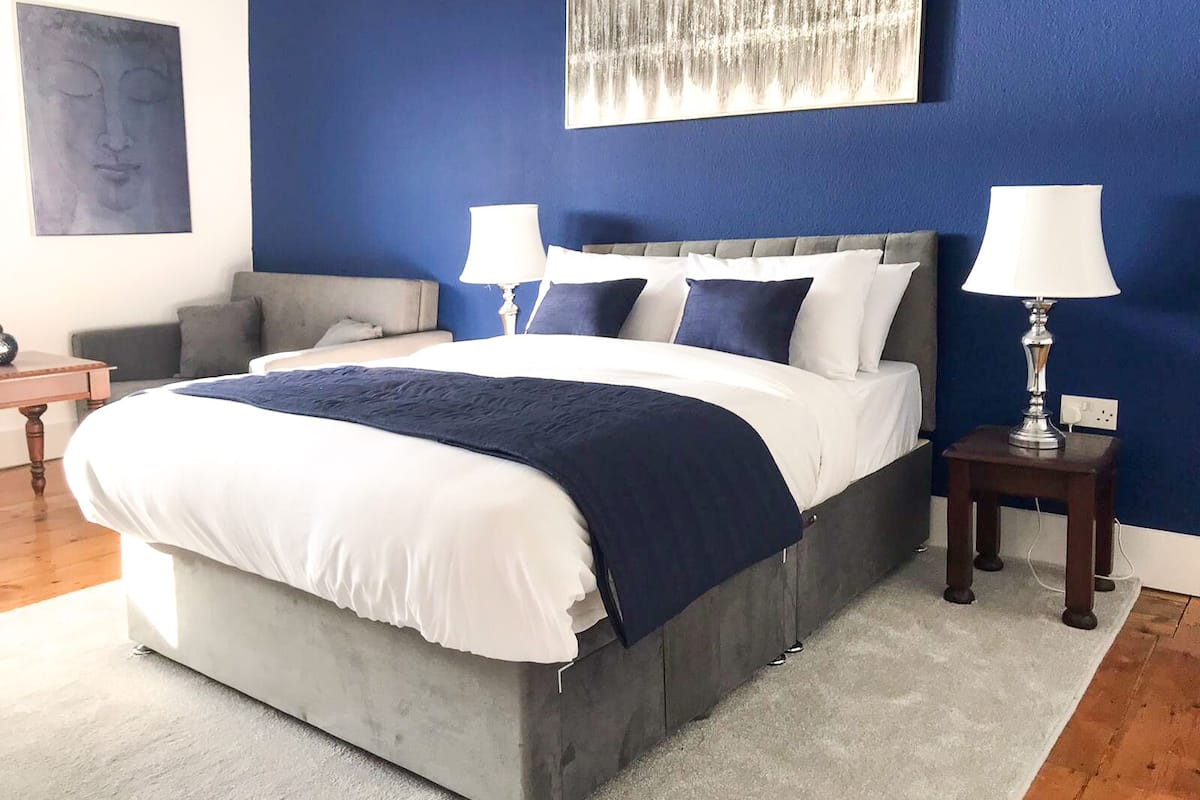Quarto com cama grande, duas mesas de cabeceira com luminárias, sofá cinza e parede azul decorativa.