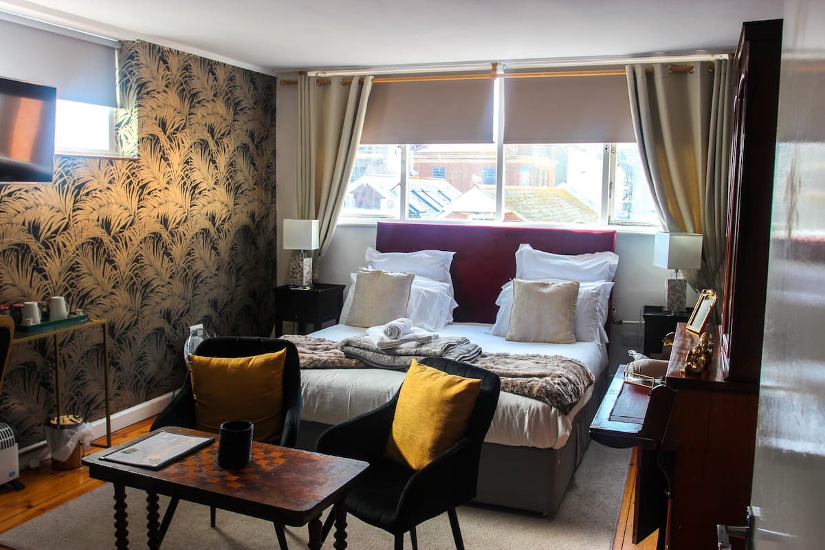 Quarto de hotel com cama, duas cadeiras, uma mesinha, janela com cortinas, televisão na parede e vista para prédios.
