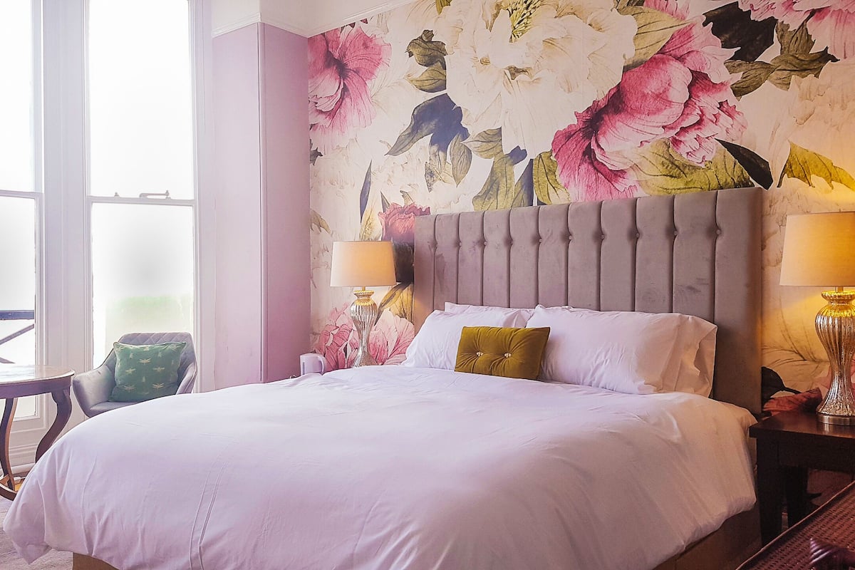 Quarto de hotel com uma cama grande, duas mesas de cabeceira com abajures, uma cadeira e papel de parede floral.