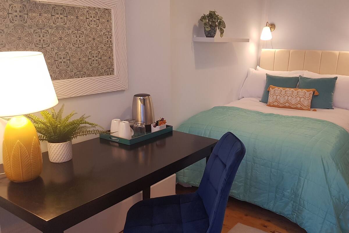Quarto de hotel bem arrumado, com cama, abajures, uma mesa com luminária, uma cadeira e uma pequena planta.