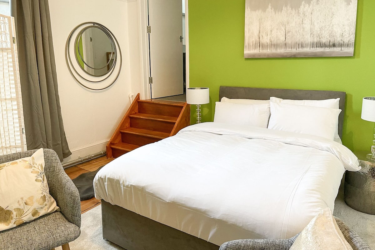 Quarto de hotel com cama, uma cadeira, escada, quadro e espelho.