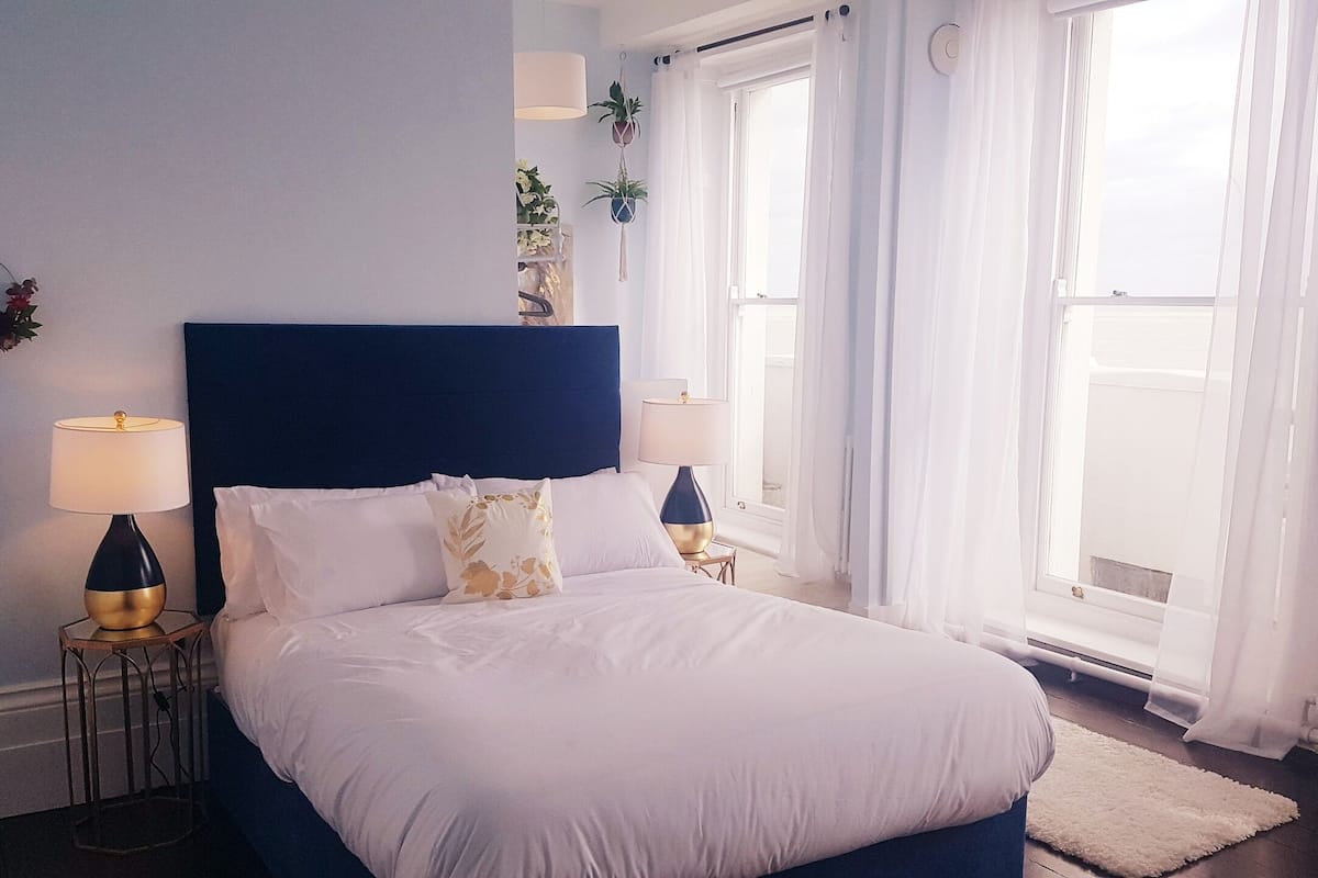 Quarto com uma cama grande, duas mesas de cabeceira, uma luminária na parede e uma janela com cortinas leves.