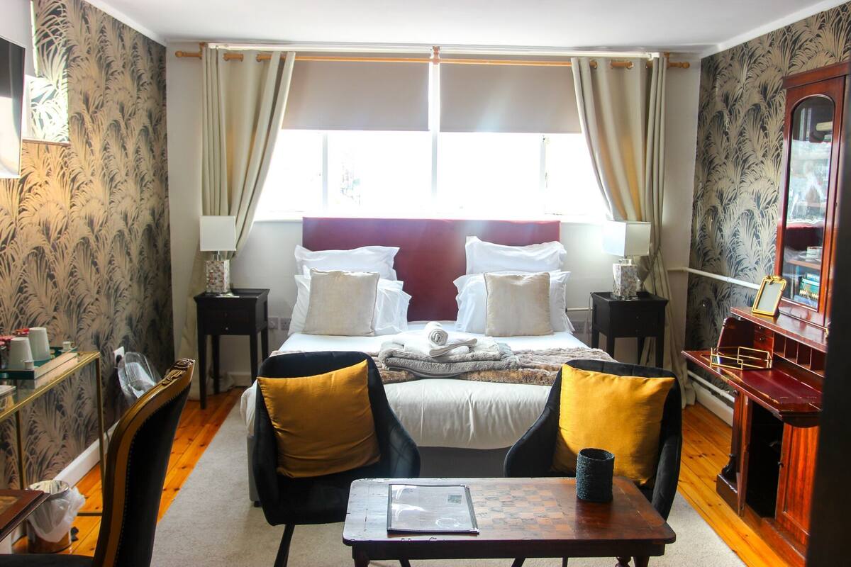 Quarto de hotel com cama, duas cadeiras, uma mesa, escrivaninha e espelho.