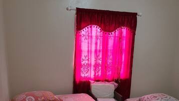 Quarto com duas camas de solteiro, cada uma com roupa de cama e almofadas estampadas com flores. Uma cortina vermelha com desenho floral cobre a janela, e uma cadeira branca está colocada contra a parede.