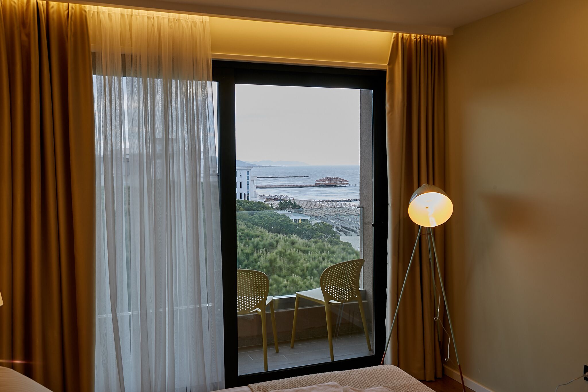 Quarto de hotel com uma janela grande, uma luminária de chão e duas cadeiras.