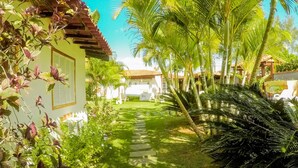 Uma trilha leva a um edifício com telhado de telhas, cercado por vegetação exuberante e palmeiras.