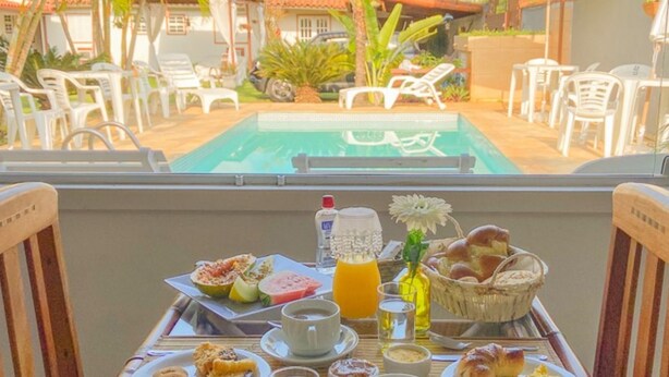 Bandeja de café da manhã com diversos alimentos e bebidas, piscina ao fundo e área externa com cadeiras brancas.