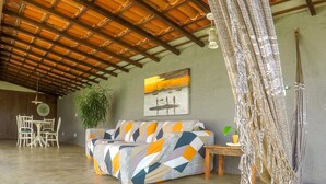 Quarto com teto de madeira, sofá com capa de padrão geométrico, pintura de um pôr do sol, planta e mesa com luminária.