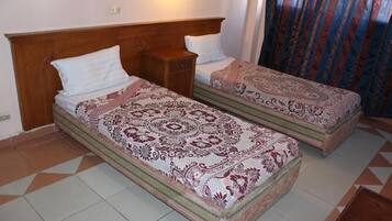 Quarto com duas camas de solteiro, cada uma com cabeceira de madeira e colcha estampada. Há uma mesinha de cabeceira entre as camas, e o chão é coberto por azulejos.