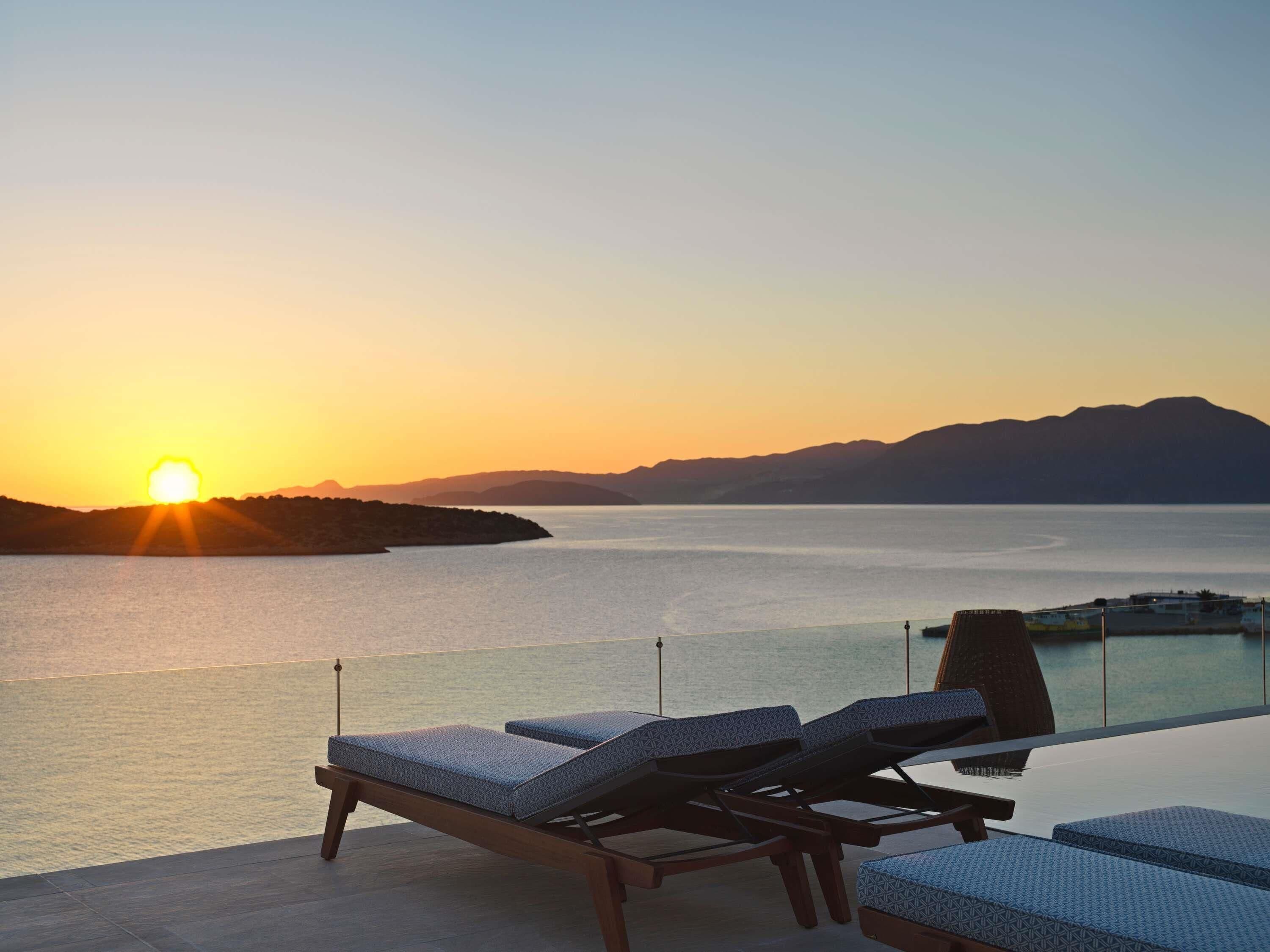 Vista do pôr do sol a partir de uma piscina infinita, com espreguiçadeiras e uma cordilheira ao longe.