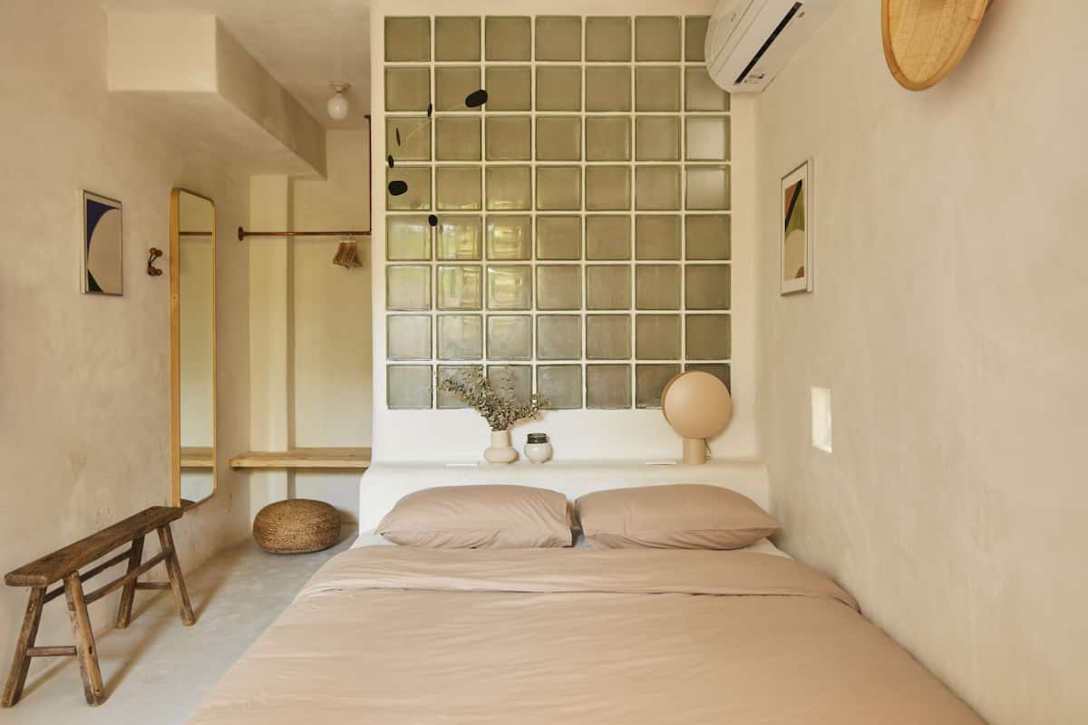 Quarto com uma janela grande, um banco de madeira, um banquinho redondo trançado e uma cama com duas almofadas.