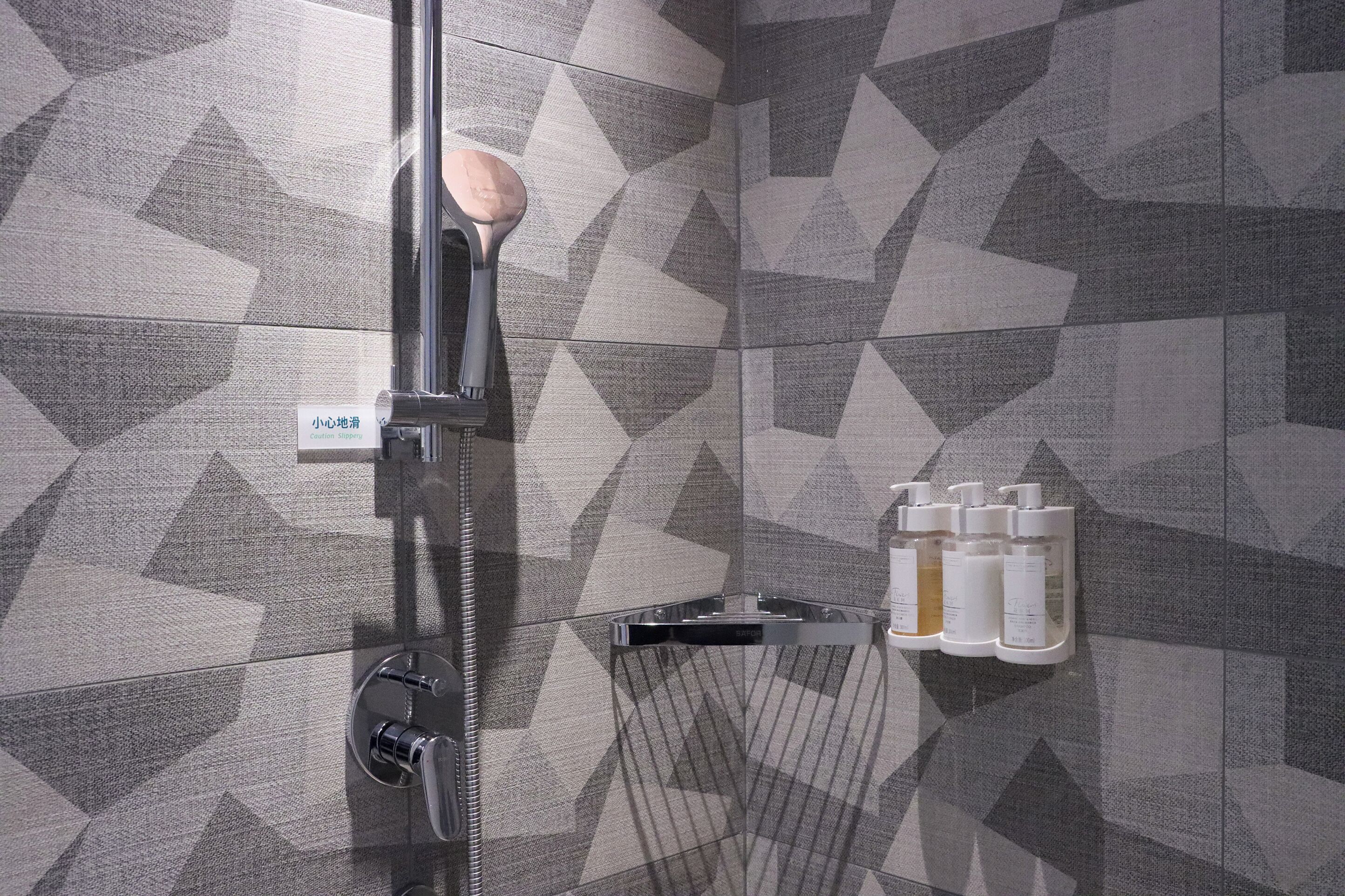 Área de chuveiro com padrão geométrico em azulejos, chuveiro manual e um conjunto de produtos de higiene pessoal na parede.