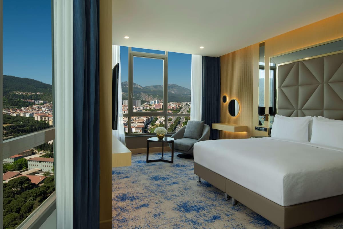 Quarto de hotel com uma cama grande, uma cadeira, uma mesa pequena e vista para a cidade através de amplas janelas.