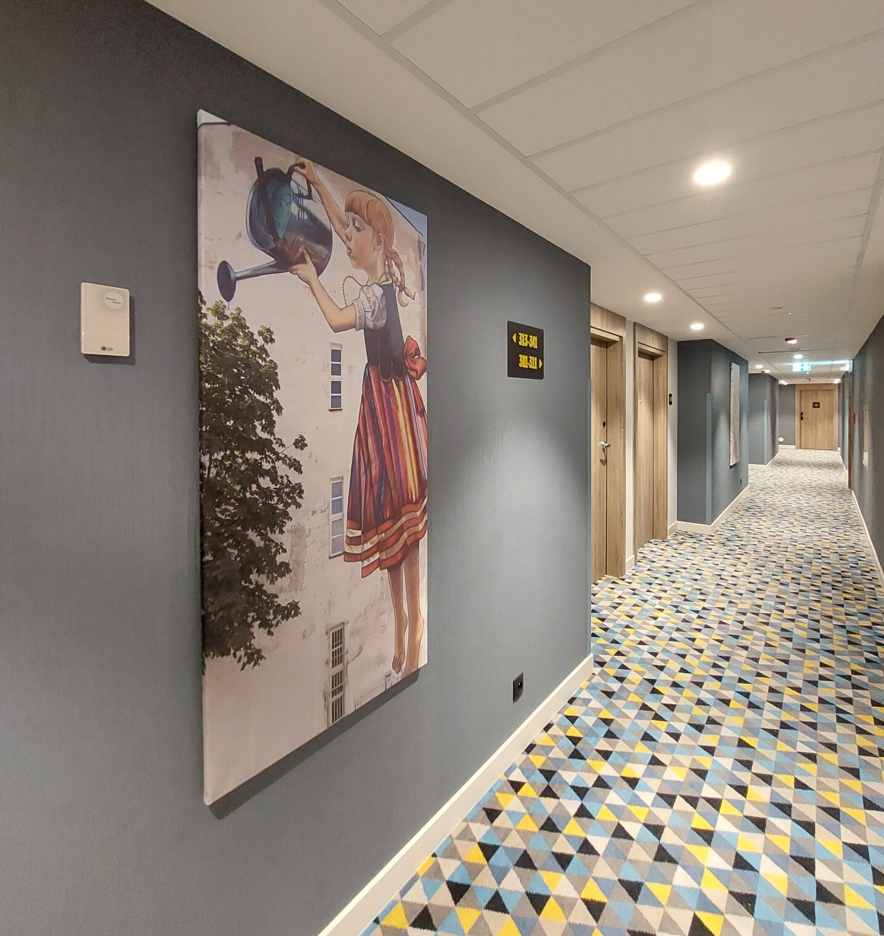 Um corredor de hotel com um piso de padrão geométrico, um grande mural na parede e placas de sinalização.