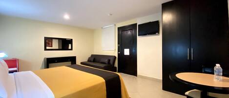 Quarto de hotel com cama, escrivaninha, cadeira, mesa pequena, televisão e guarda-roupa.