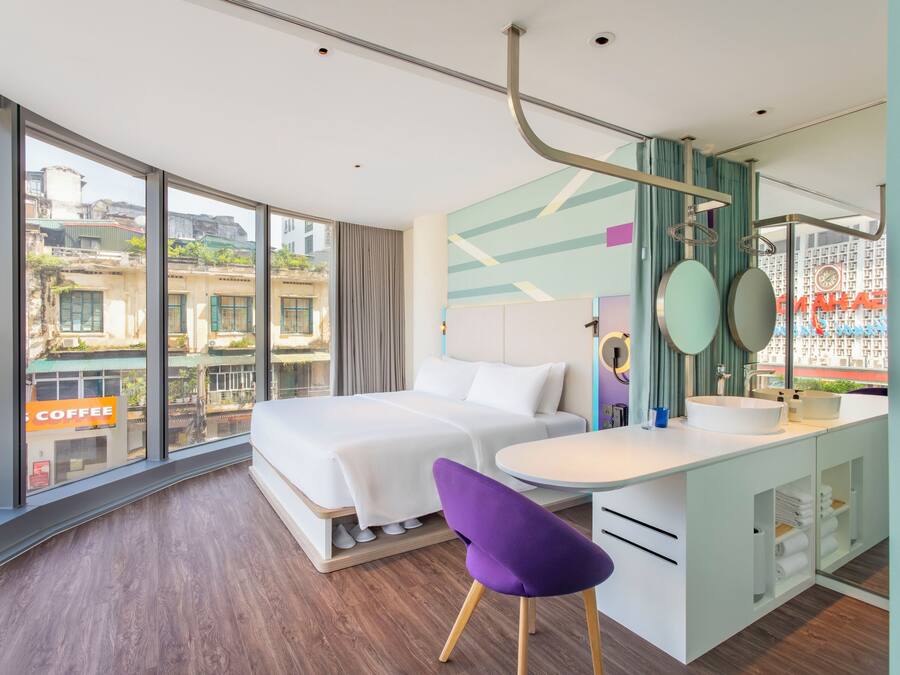 Quarto de hotel moderno com uma cama grande, uma escrivaninha e uma pia, e vista da cidade através de grandes janelas.