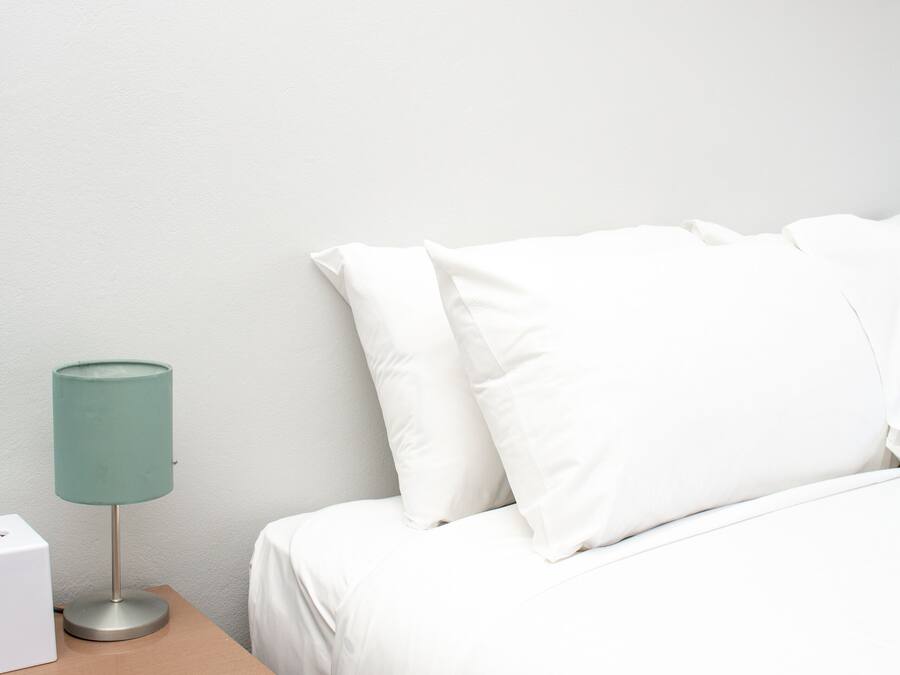 Uma cama bem arrumada com roupa de cama branca, um criado-mudo de madeira com abajur azul-turquesa e uma parede branca.