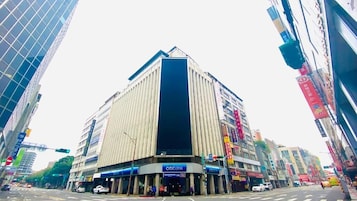 Um edifício moderno com uma característica vertical marcante, provavelmente um banco ou sede corporativa, rodeado por outros edifícios comerciais e ruas movimentadas.