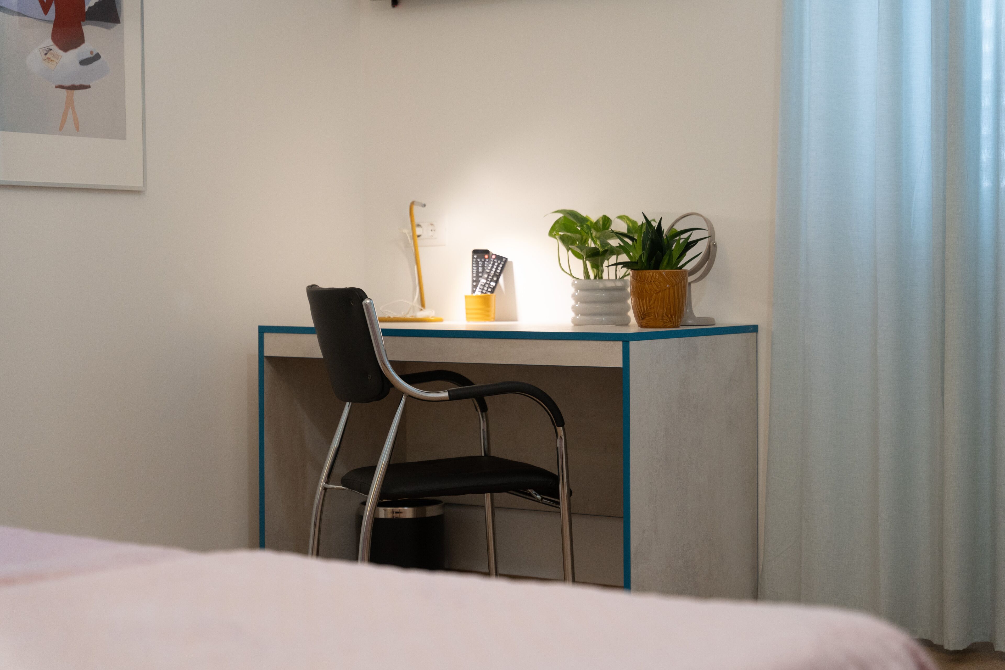 Quarto de hotel bem arrumado com uma cama, uma escrivaninha com cadeira, um vaso de planta, um controle remoto e um quadro na parede.