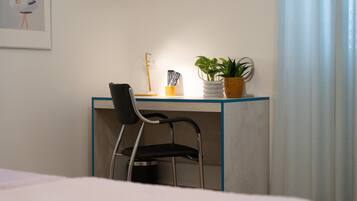 Quarto de hotel bem arrumado com uma cama, uma escrivaninha com cadeira, um vaso de planta, um controle remoto e um quadro na parede.