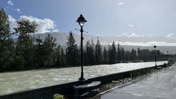 Uma passarela às margens do rio, com bancos e postes de luz, com vista para o rio e uma área arborizada com montanhas ao fundo.