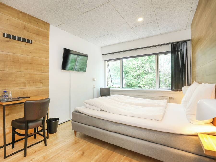 Quarto com cama, escrivaninha, cadeira, televisão e janela com vista para árvores.