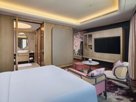 Quarto de hotel moderno com uma cama grande, um espaço de estar com uma cadeira e mesa, uma televisão e um banheiro com espelho e pia.
