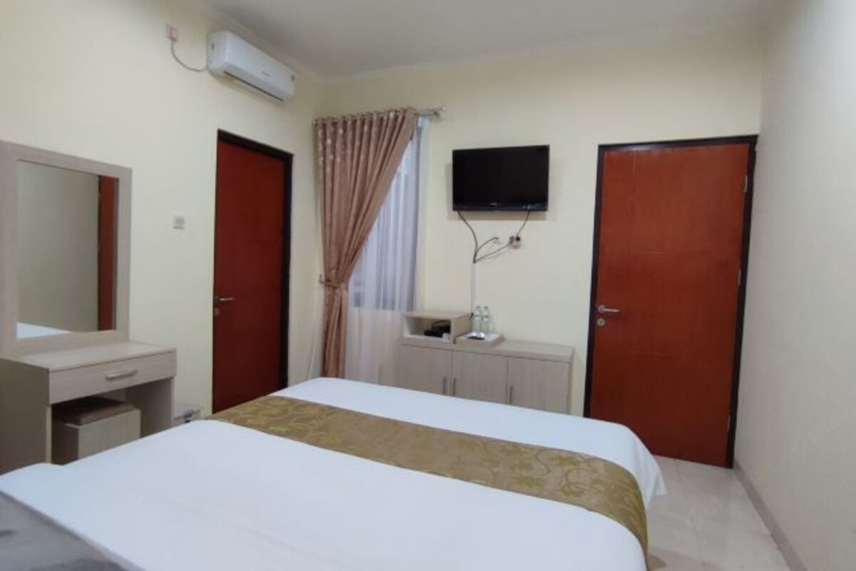 Quarto de hotel com cama, escrivaninha, espelho, televisão e duas portas.