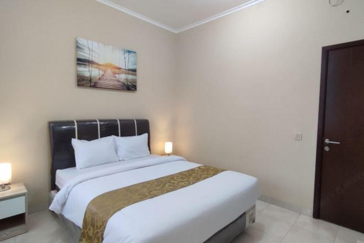 Cama bem arrumada com lençóis brancos e detalhes dourados, um abajur e um quadro na parede.