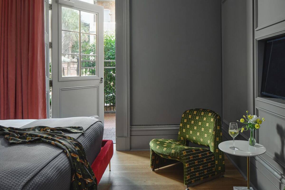 Quarto de hotel com cama, uma cadeira, mesinha com copo e flores, e vista para o exterior através de uma porta.