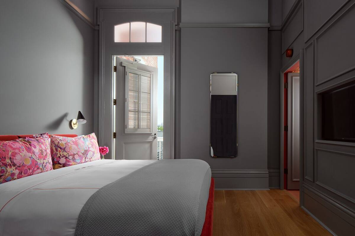 Quarto com cama, janela, porta e televisão presa na parede.