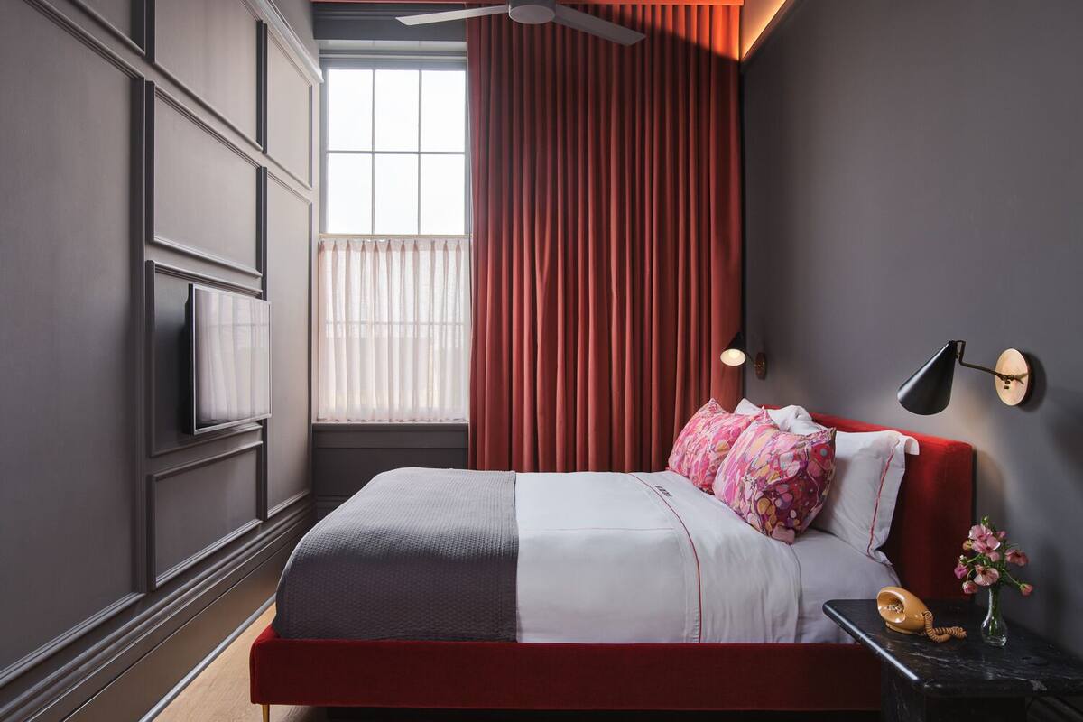 Quarto moderno com cama grande, ventoinha no teto, janela com cortinas, mesa de cabeceira com luminária e um vaso com flores.