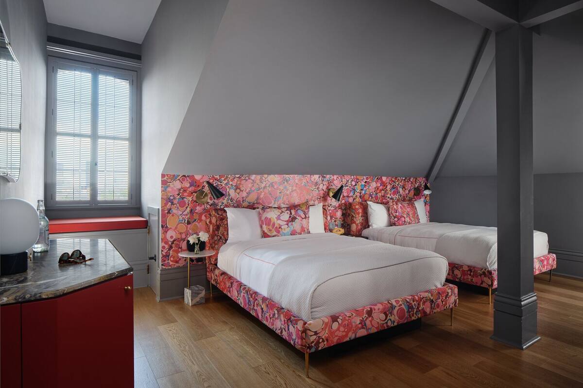 Quarto com teto inclinado, uma cama grande com cabeceira estampada com motivos florais, uma mesinha de cabeceira e uma janela com persianas.