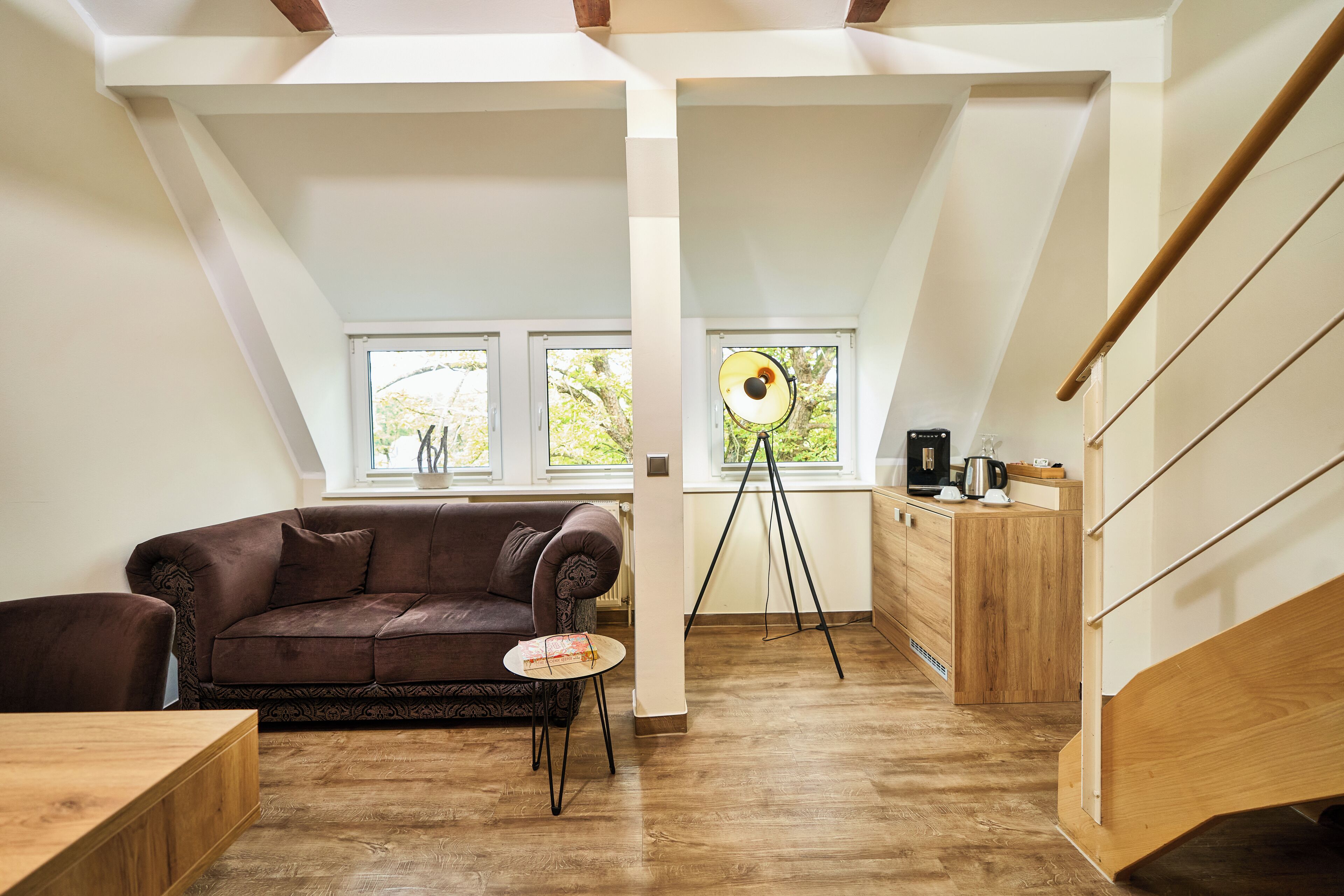 Quarto estilo loft com sofá de couro marrom, uma pequena mesa redonda e uma luminária de tripé. Uma escada de madeira à direita e uma área de cozinha com cafeteira e armários.