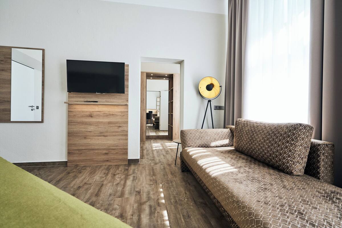 Quarto de hotel moderno com piso de madeira, um sofá com almofadas estampadas, uma TV de tela plana fixada em um painel de madeira, um abajur e um espelho grande.