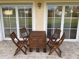 Um pátio com mesa de madeira e quatro cadeiras, grandes portas de vidro e chão de tijolos.