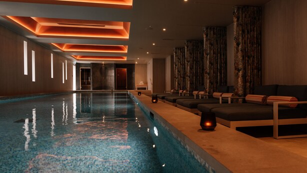 Piscina interior moderna, com design alongado e estreito, rodeada por áreas de estar com cortinas estampadas e um sofá.