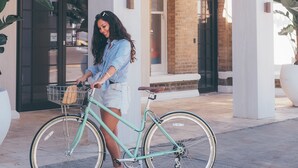 Uma pessoa ao lado de uma bicicleta azul-turquesa com cesto, em frente a um edifício com uma placa que diz "THE NICI".