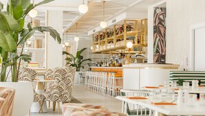 Interior moderno de restaurante com mobiliário branco e castanho estampado, área de bar e uma planta grande.