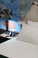 Quarto de hotel com cama, mesa de cabeceira com telefone e luminária, e parede com um mural.